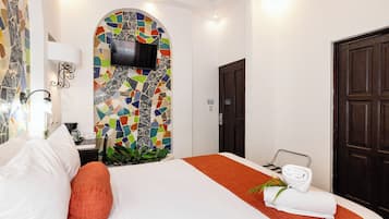 Quarto de hotel com uma cama grande, televisão presa a uma parede com mosaico colorido, porta de madeira escura e uma pequena planta na mesinha de cabeceira.