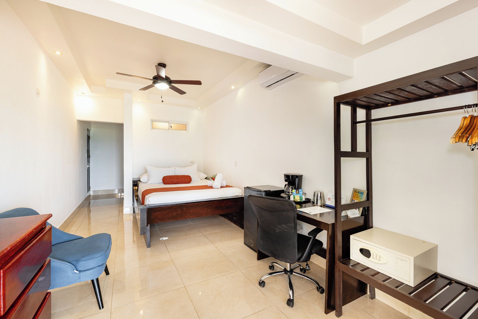 Quarto de hotel com cama, escrivaninha, cadeira, guarda-roupa e ventoinha no teto.