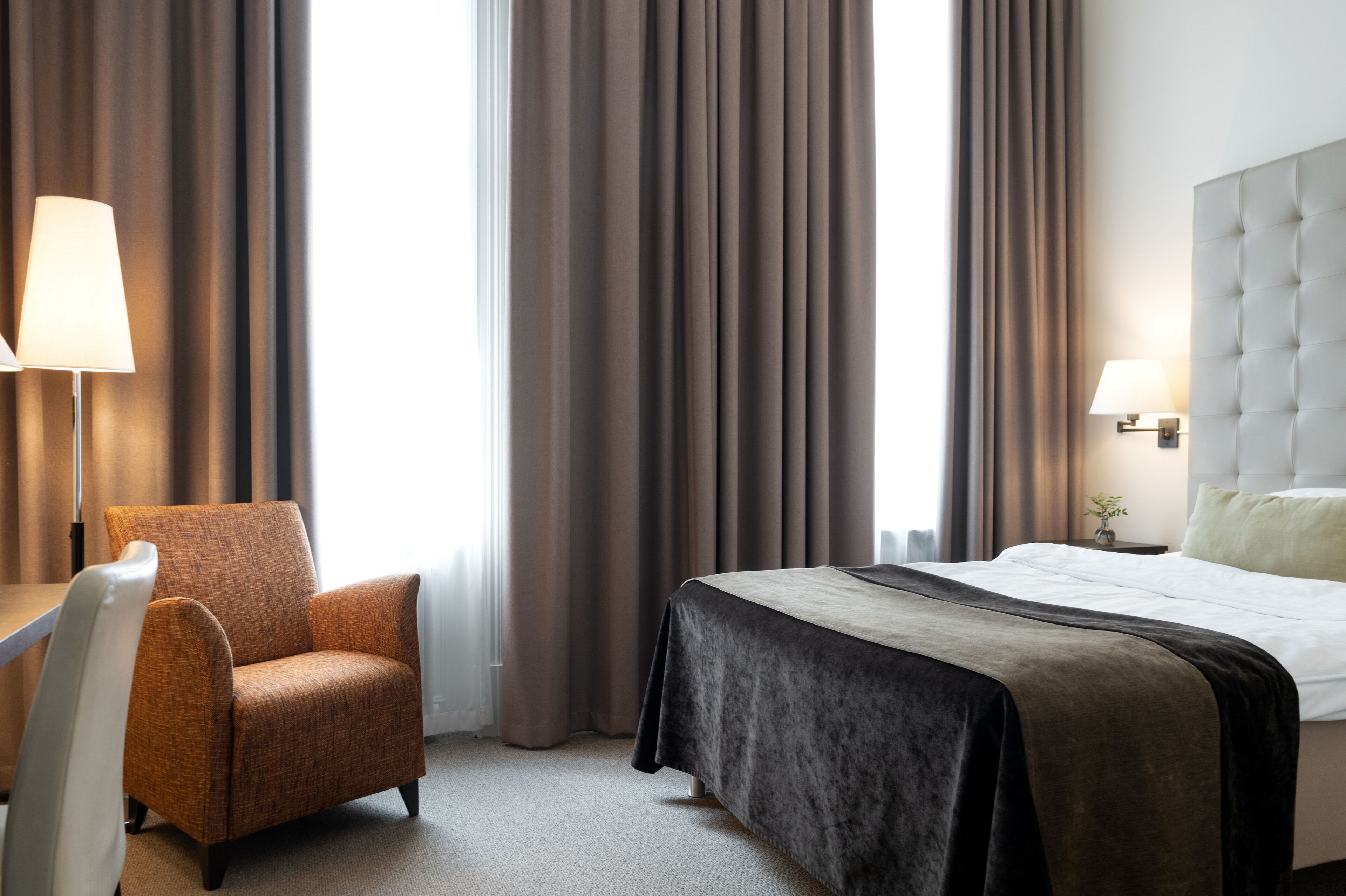 Quarto de hotel com cama, cadeira, abajur e cortinas.