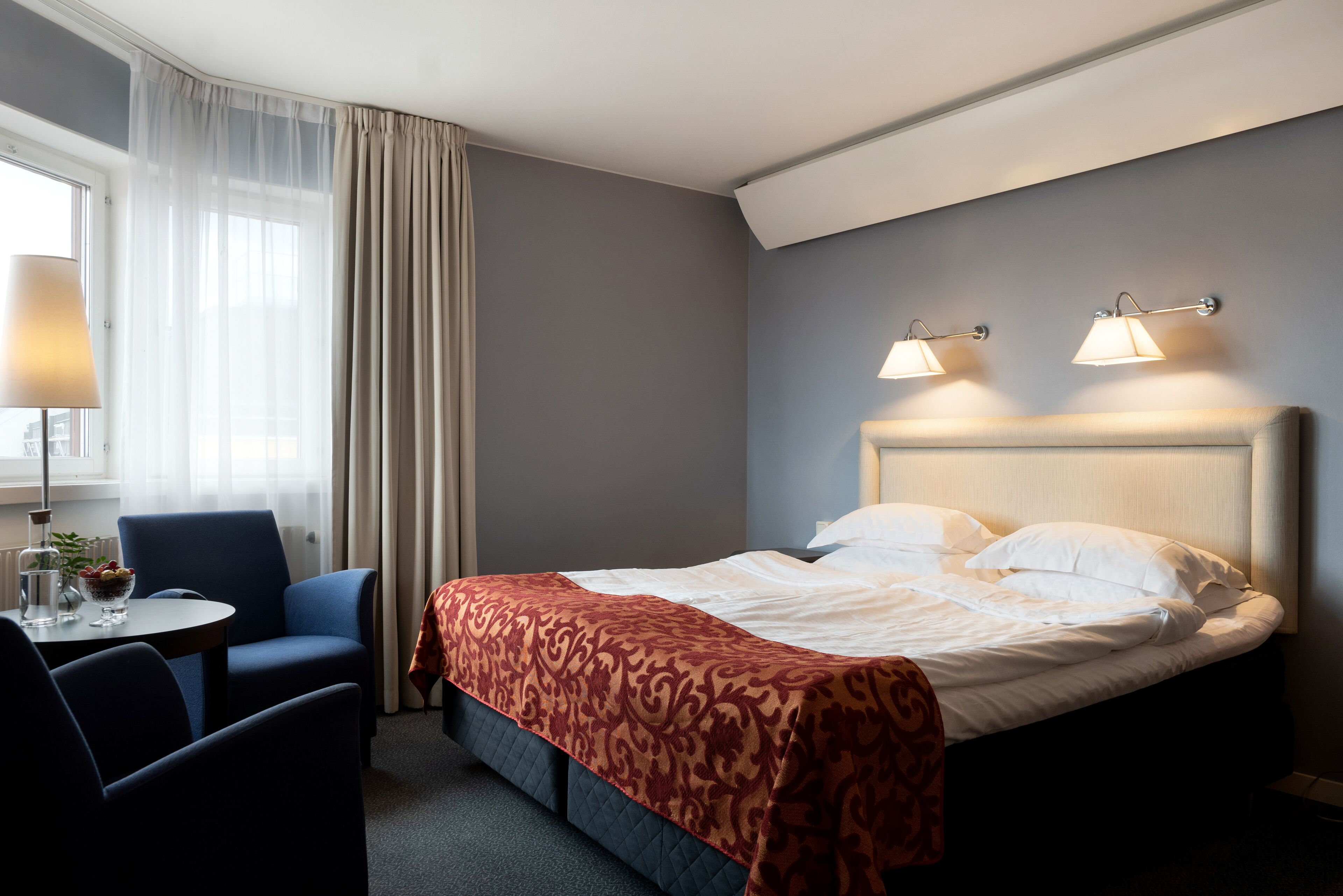 Quarto de hotel com cama, escrivaninha, cadeira, abajur e janela com cortinas.