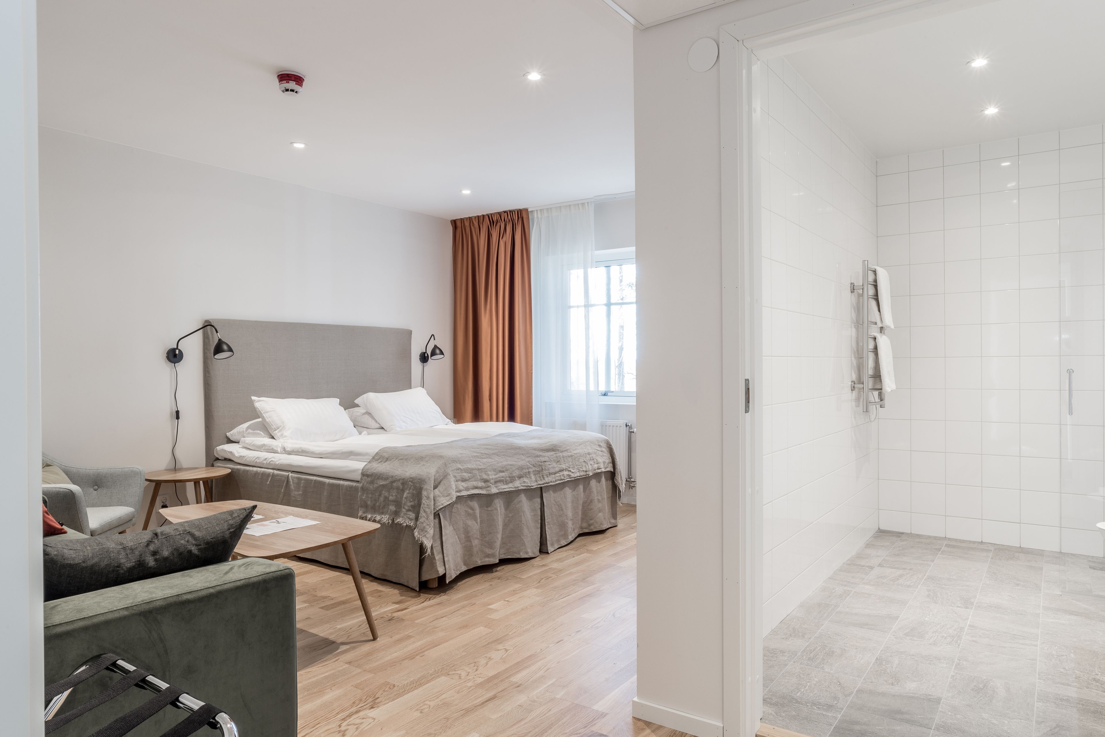 Quarto de hotel moderno com cama, escrivaninha, cadeira e área de chuveiro.