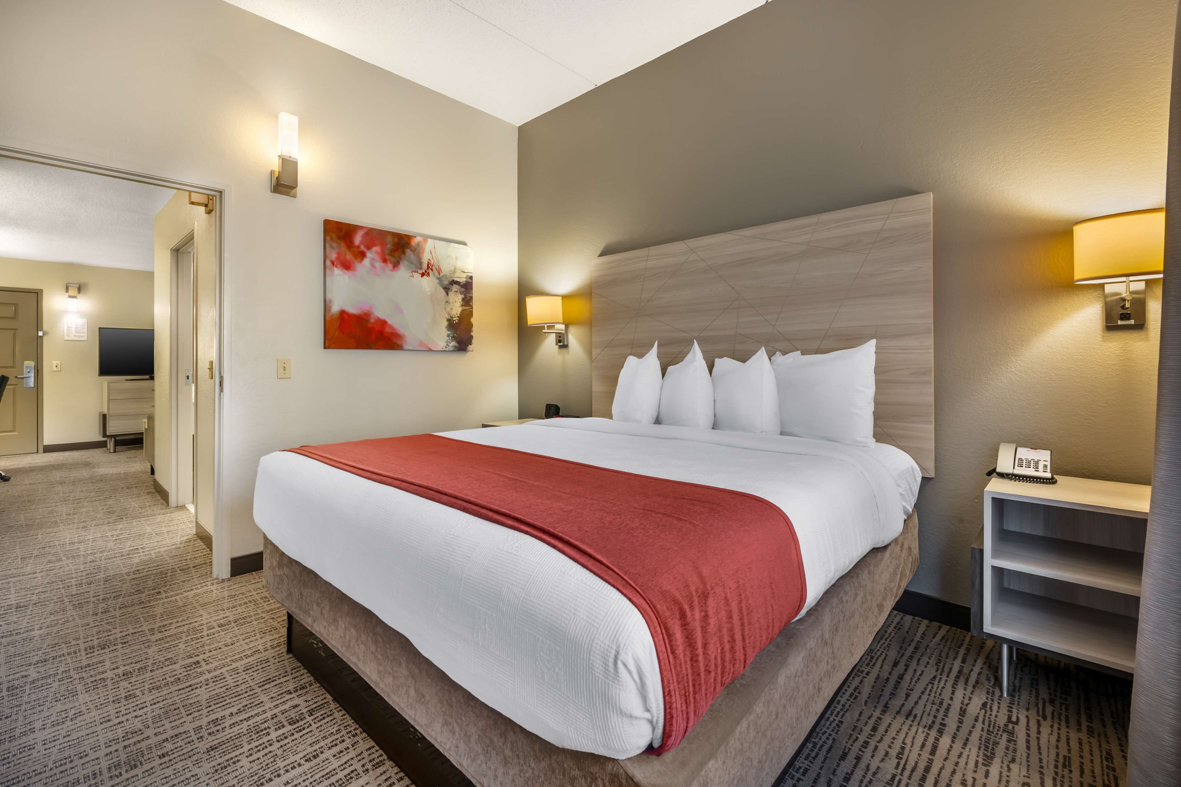 Quarto de hotel com uma cama grande, manta vermelha, cabeceira com padrão geométrico, quadro na parede e escrivaninha com luminária e telefone.