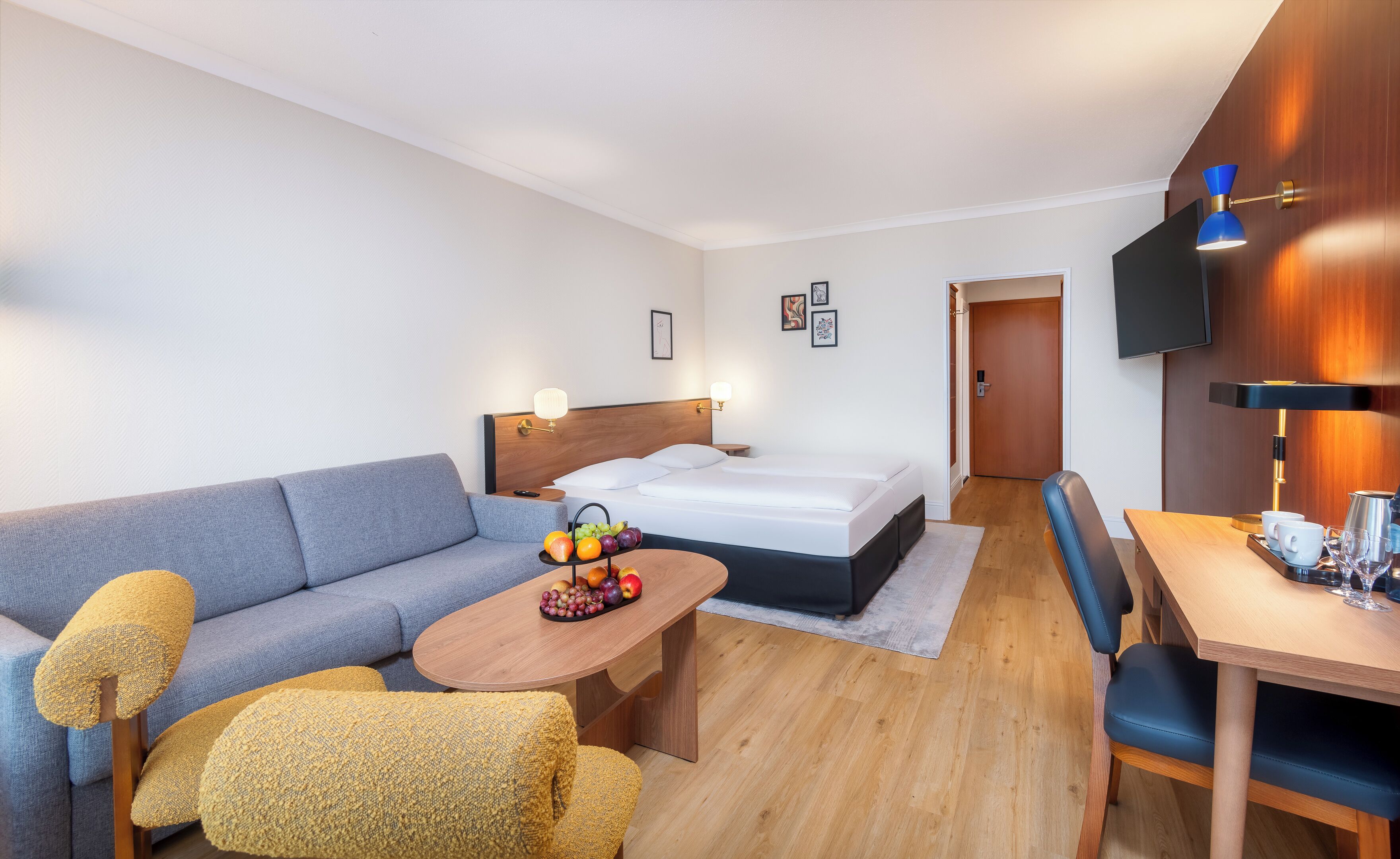 Quarto de hotel moderno com sofá, mesa com frutas, cama, escrivaninha e televisão na parede.