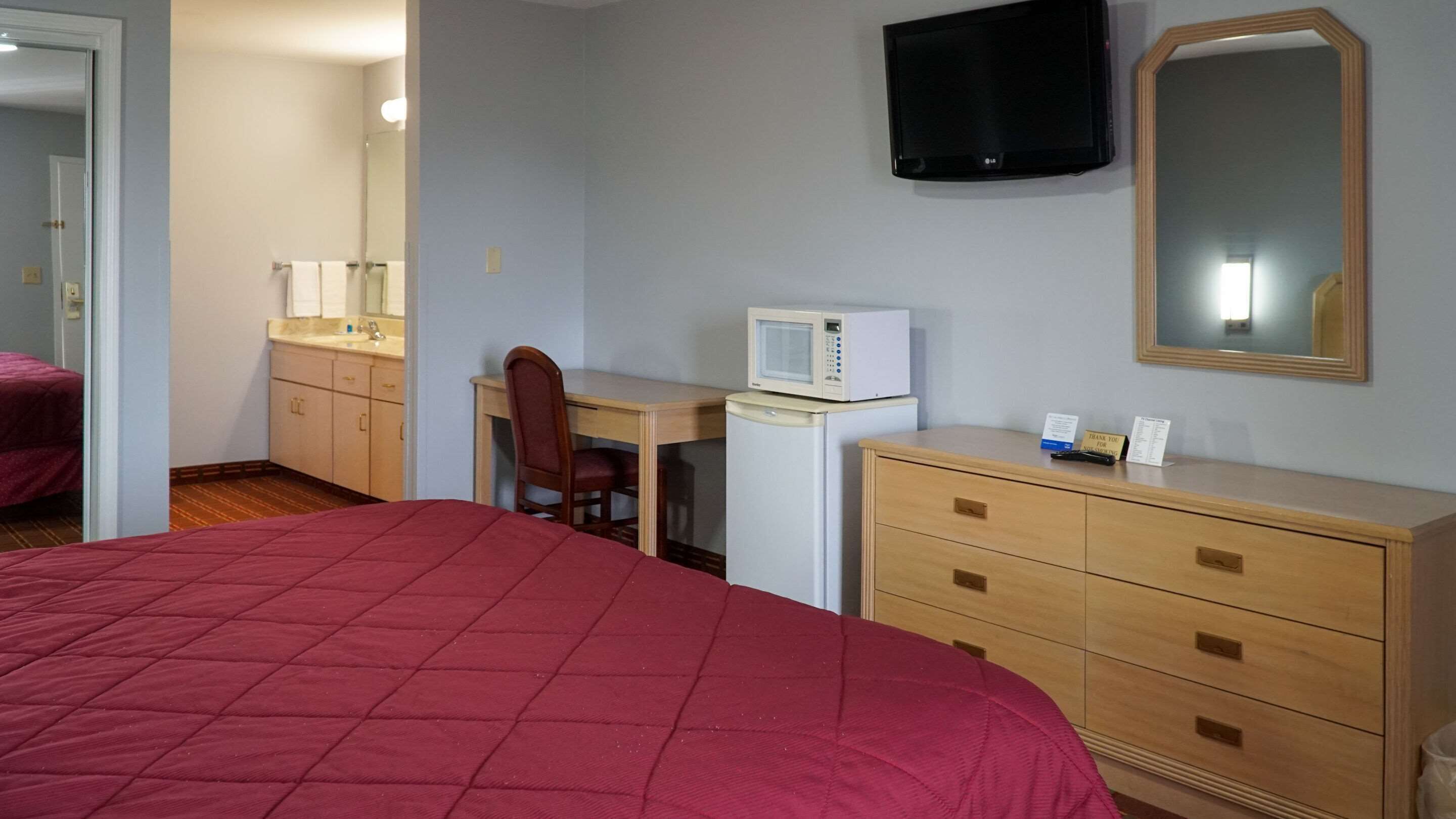 Quarto de hotel com cama, escrivaninha com cadeira, micro-ondas, cômoda, televisão e espelho.