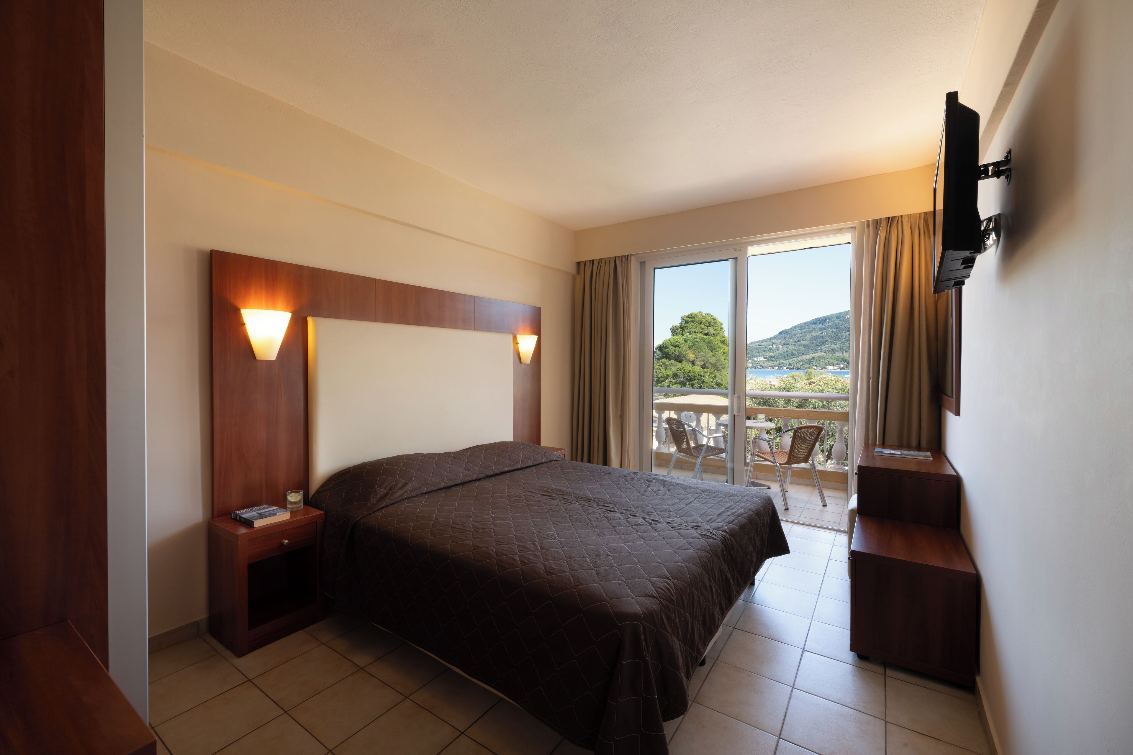 Quarto de hotel com uma cama grande, mesa de cabeceira, TV na parede e varanda com vista para o exterior.