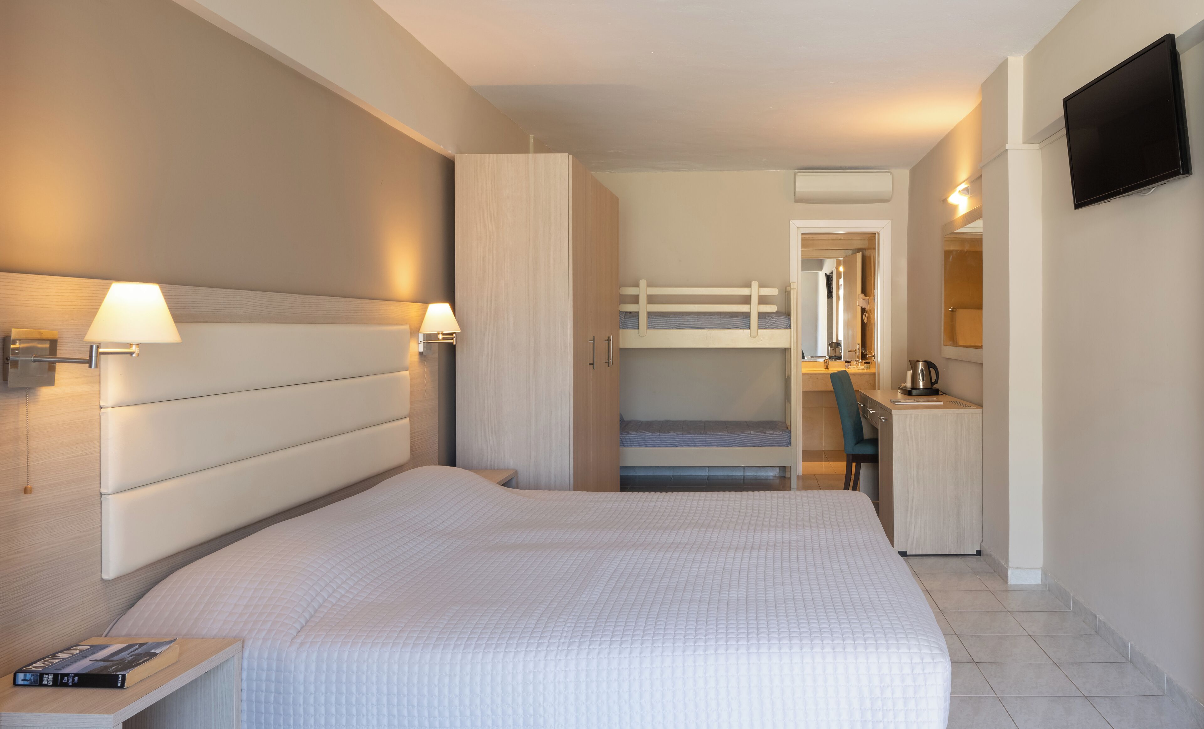 Quarto de hotel com uma cama grande, uma escrivaninha, uma beliche e um banheiro visível através de uma porta aberta.