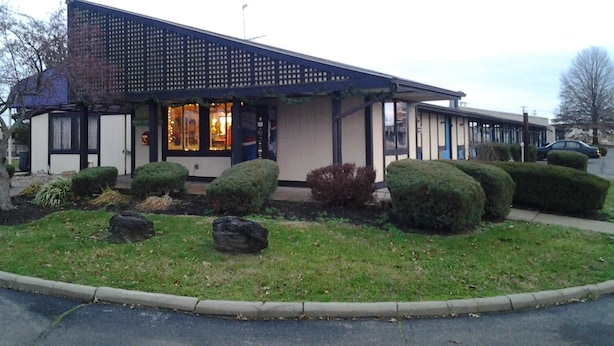 Um edifício com telhado inclinado, uma pequena varanda e um gramado bem cuidado com arbustos e pedras.