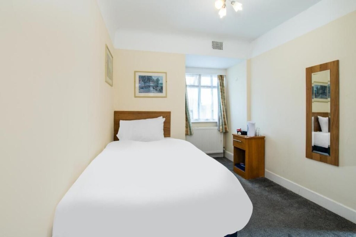 Quarto de hotel com uma cama grande, uma mesa de cabeceira de madeira, um espelho e uma janela com cortinas.