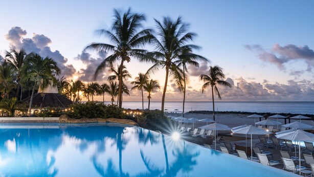 Vista da piscina com espreguiçadeiras e guarda-sóis, palmeiras e um pôr do sol sobre o oceano.