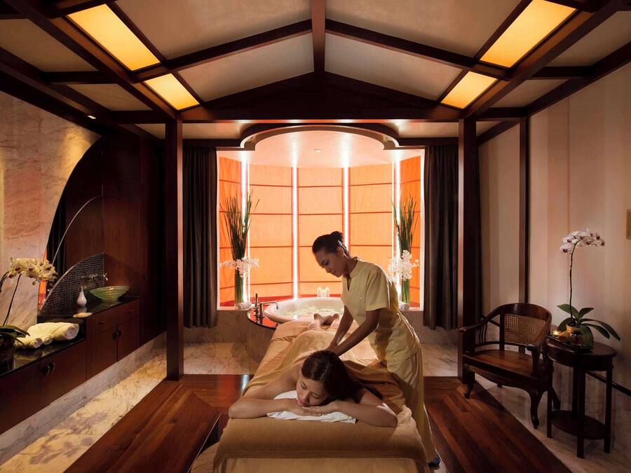 Sala de massagem com piso de madeira, uma mesa de massagem, uma cadeira e uma janela com cortinas.