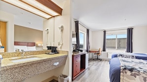 Quarto de hotel com pia de bancada de granito, espelho, secador de cabelo, escrivaninha com cadeira, cama com manta estampada e janela com cortinas.