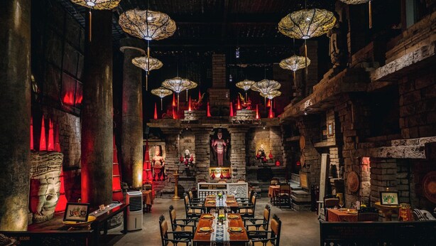 Restaurante com pouca luz, paredes de pedra, luminárias pendentes e um altar central com estátuas. Mesas preparadas para refeições.