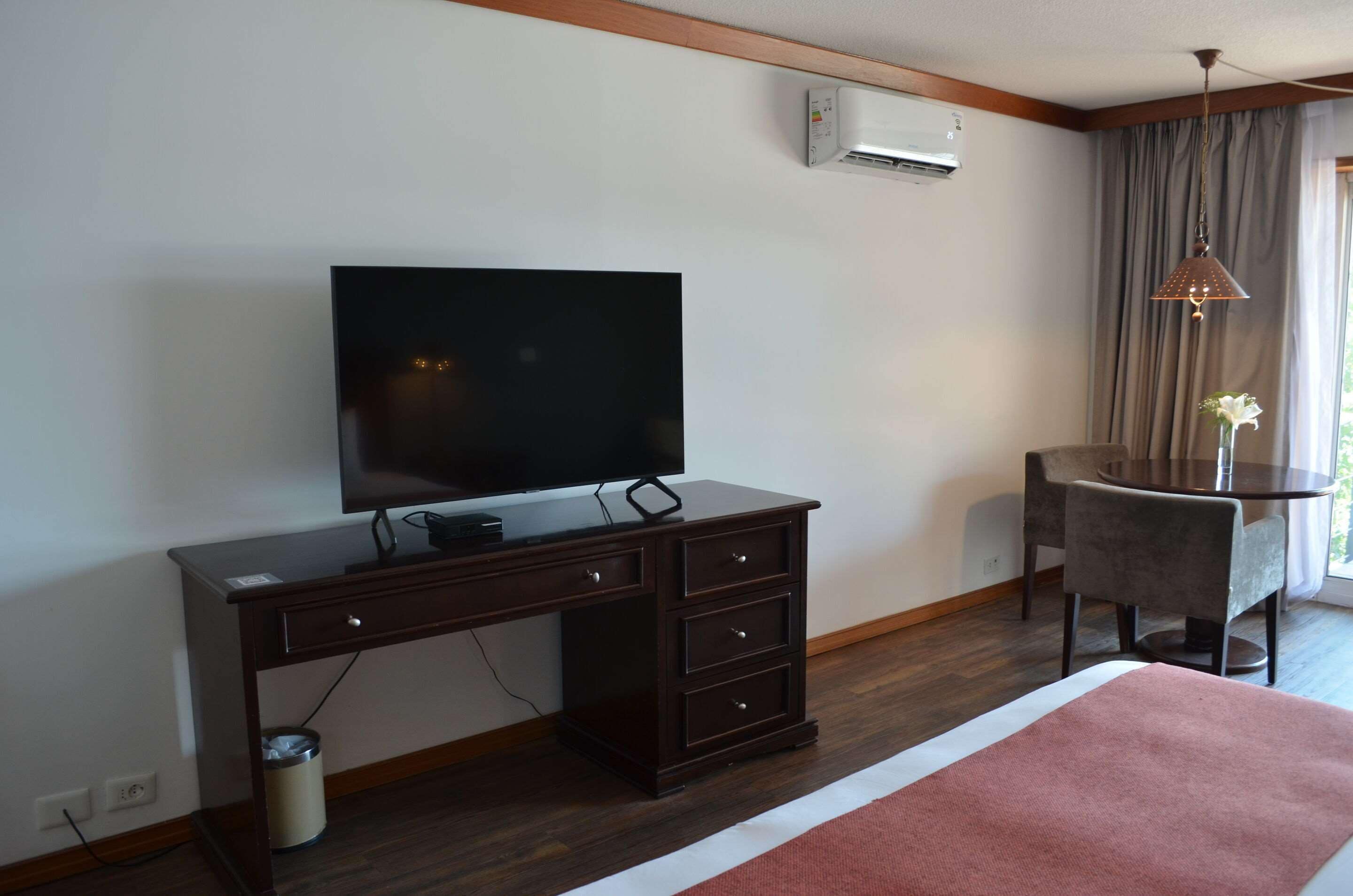 Quarto de hotel com uma televisão de tela plana grande fixada em uma cômoda de madeira, uma mesinha com duas cadeiras e uma janela com cortinas.