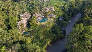 Um resort tropical com várias construções em estilo bangalô, uma piscina central e um rio que corta a propriedade.