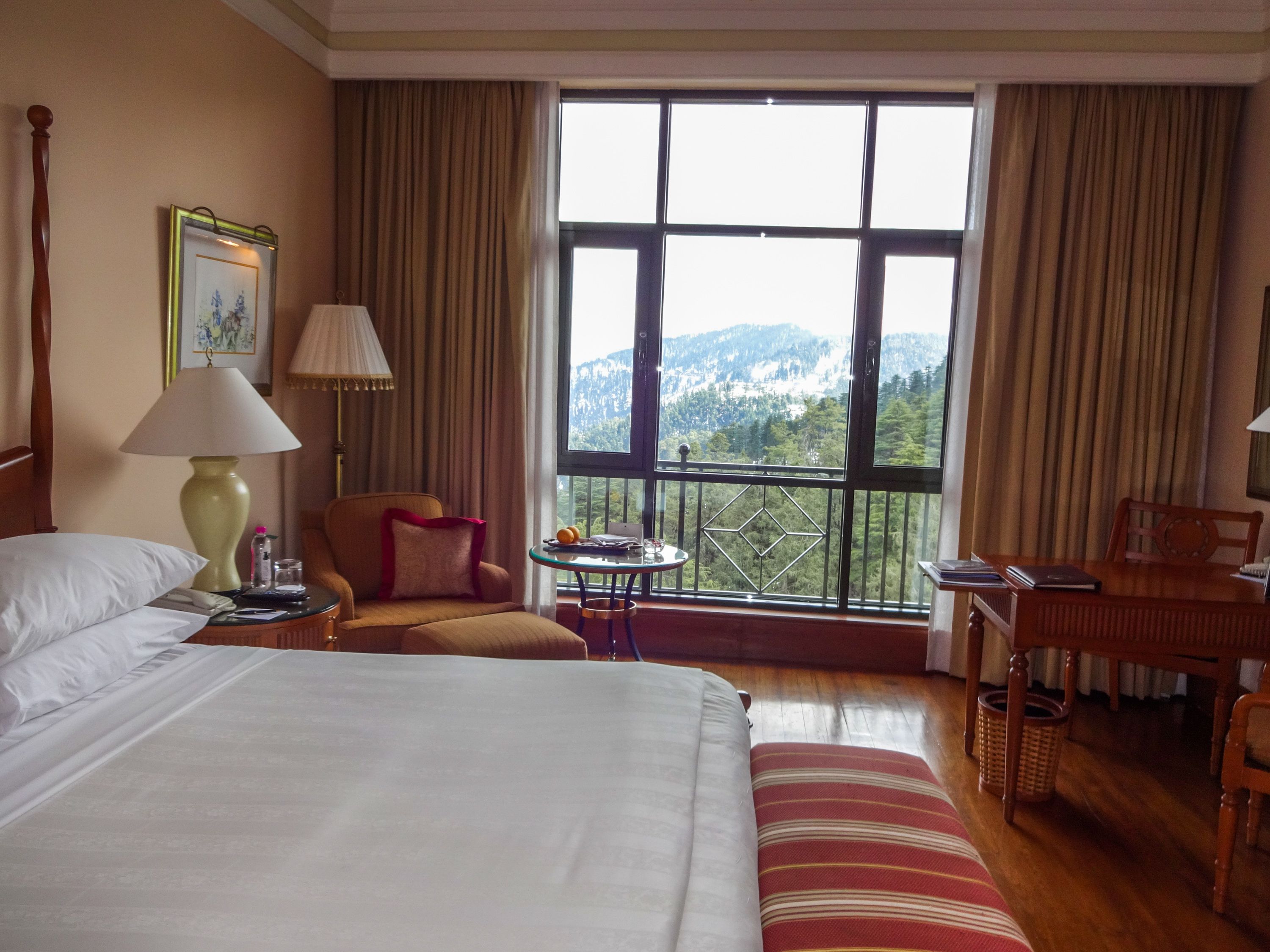 Quarto de hotel com uma cama grande, uma escrivaninha, uma cadeira, uma mesa e vista para uma paisagem montanhosa pela janela.