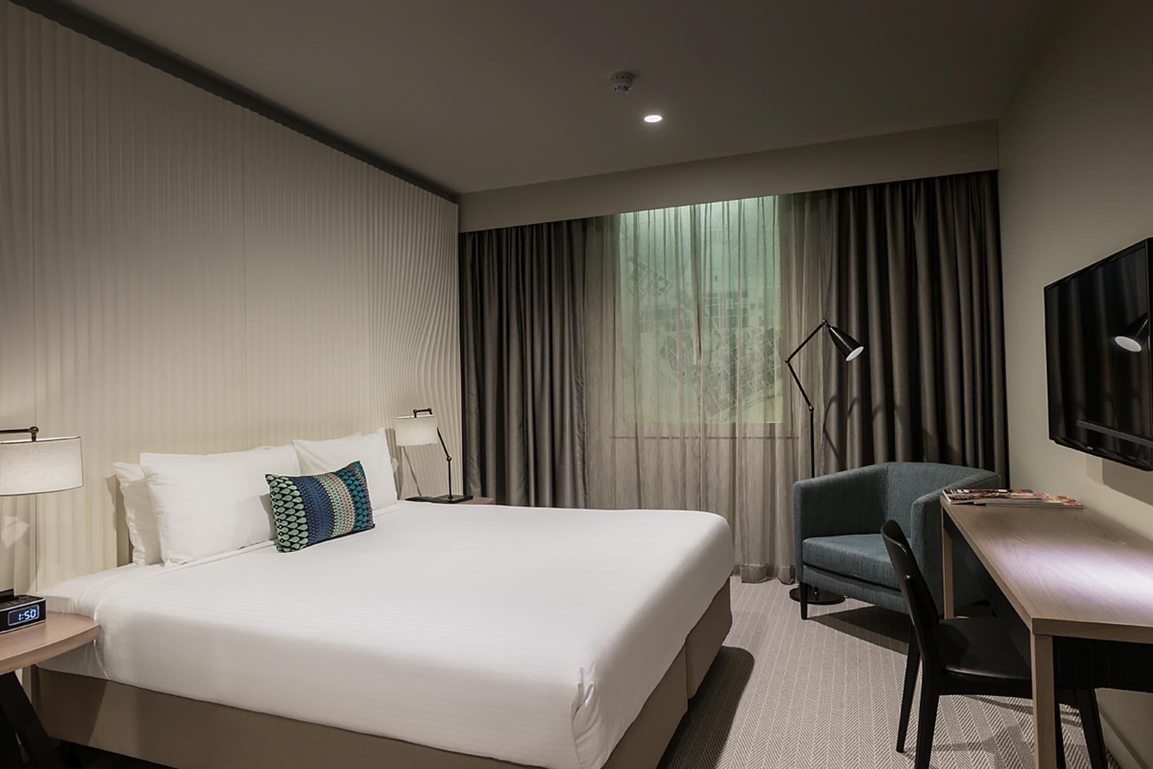 Quarto de hotel com uma cama grande, escrivaninha, cadeira, televisão e janela com cortinas.