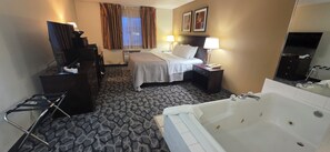 Quarto de hotel com cama, abajures, escrivaninha, cadeira, luminária de mesa, telefone, espelho, banheira e janela com cortinas.