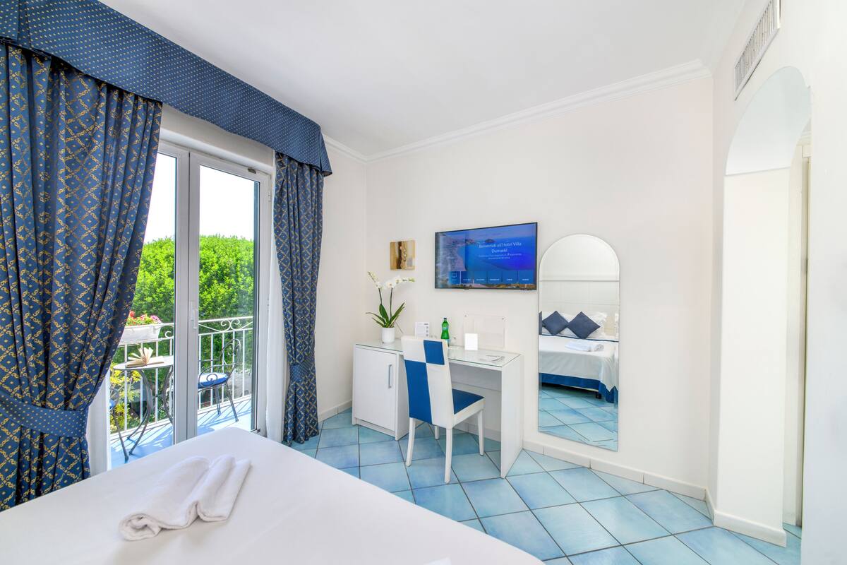 Quarto de hotel com uma cama, uma escrivaninha com cadeira, uma mesinha com planta em vaso, televisão na parede e varanda com vista para o verde.