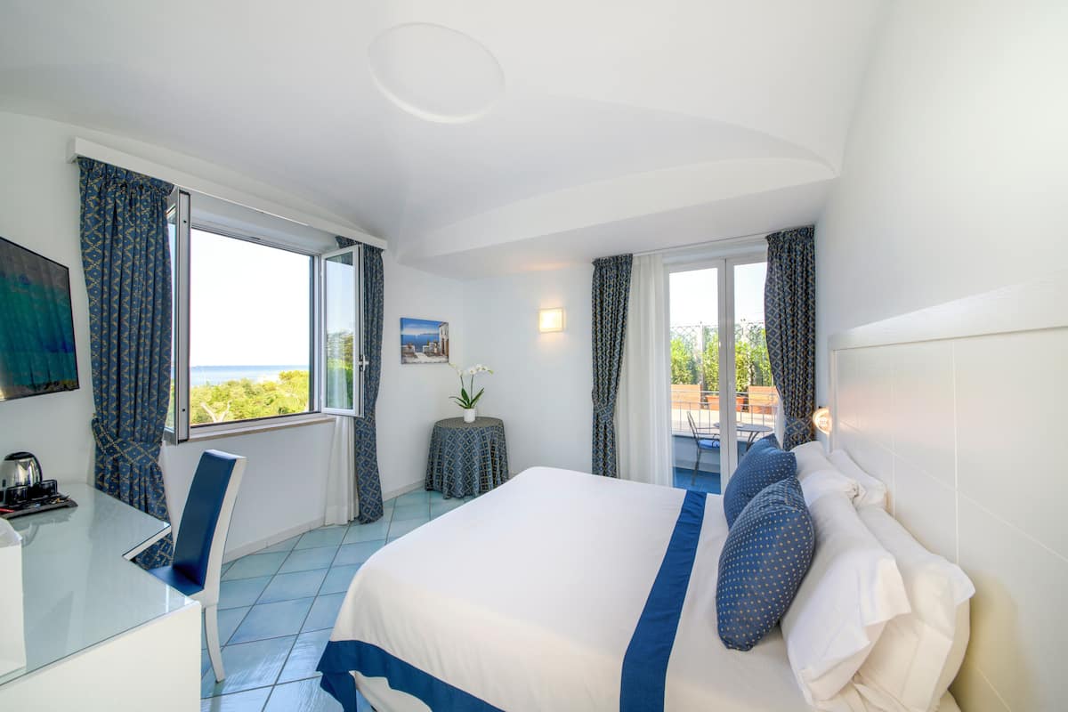 Quarto de hotel com cama, escrivaninha e cadeira. Há uma janela com cortinas, uma pequena mesa com uma planta e vista para o exterior.