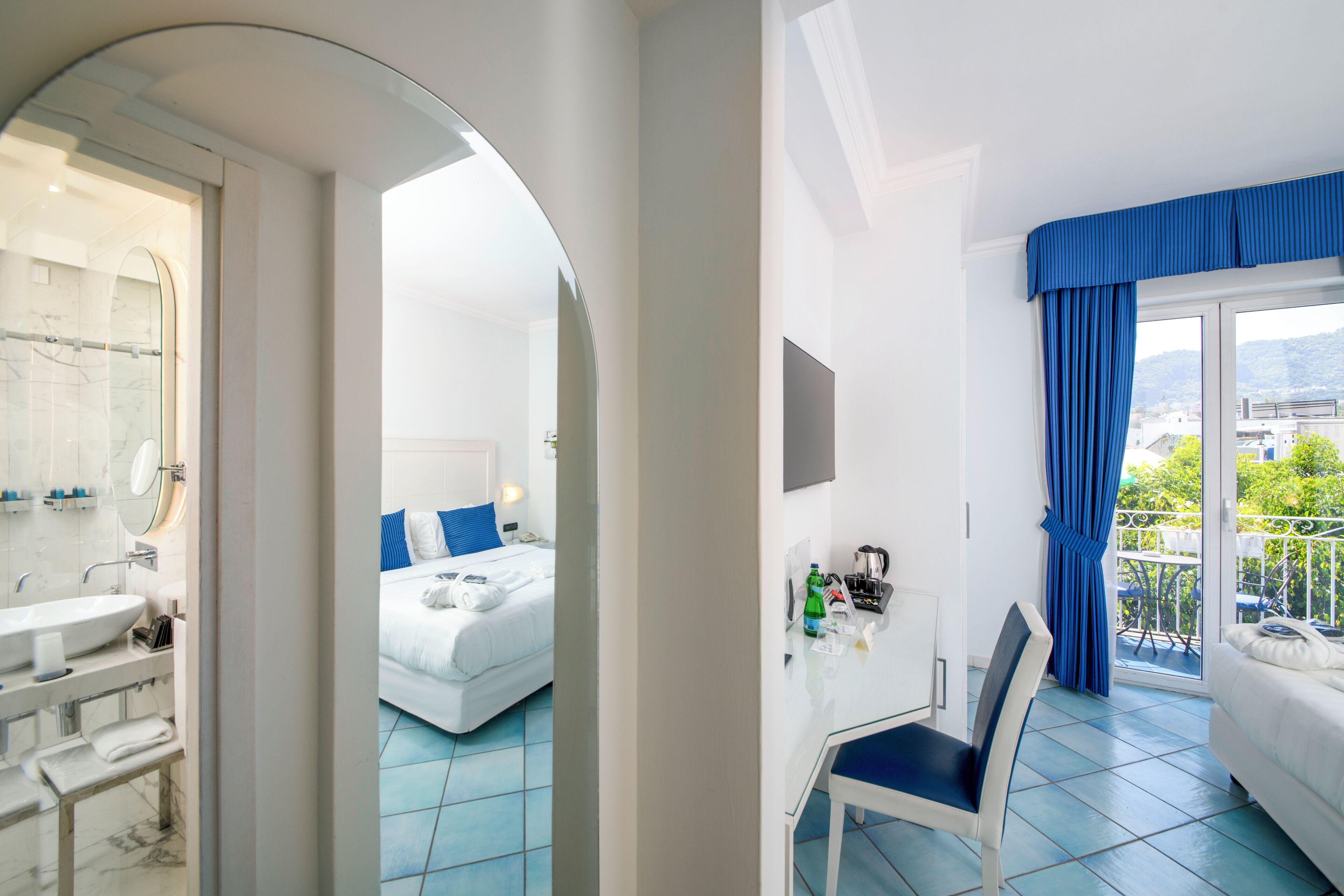 Quarto de hotel com banheiro, cama com almofadas azuis, escrivaninha com cadeira e varanda com cortinas azuis.