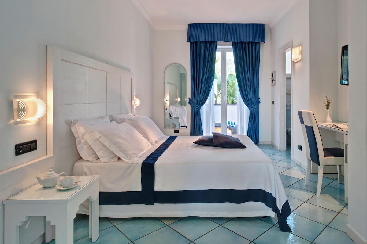 Quarto de hotel com cama, mesa de cabeceira com luminária, escrivaninha com cadeira e janela com cortinas.