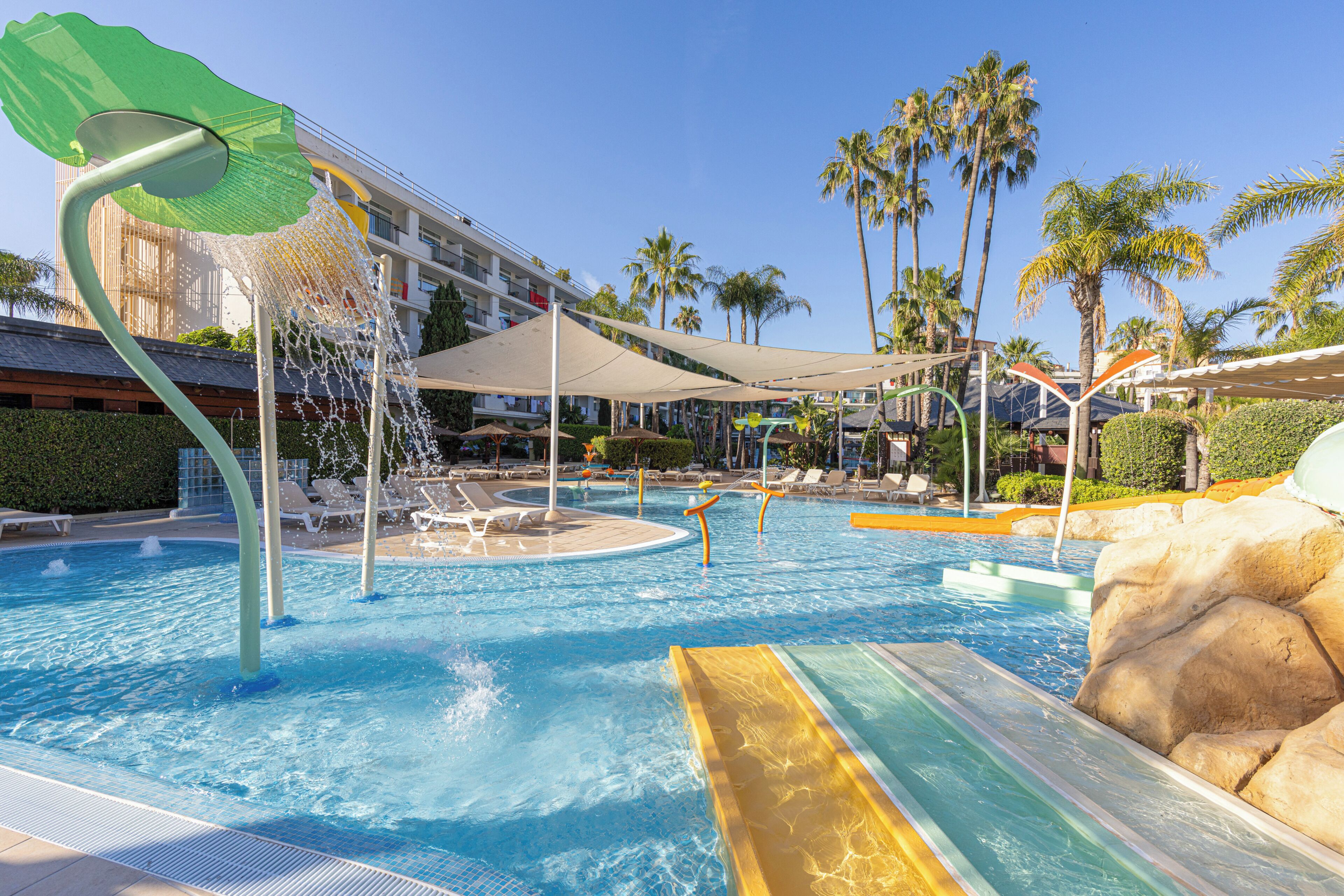 Uma piscina com escorregador, espreguiçadeiras, palmeiras e um edifício ao fundo.
