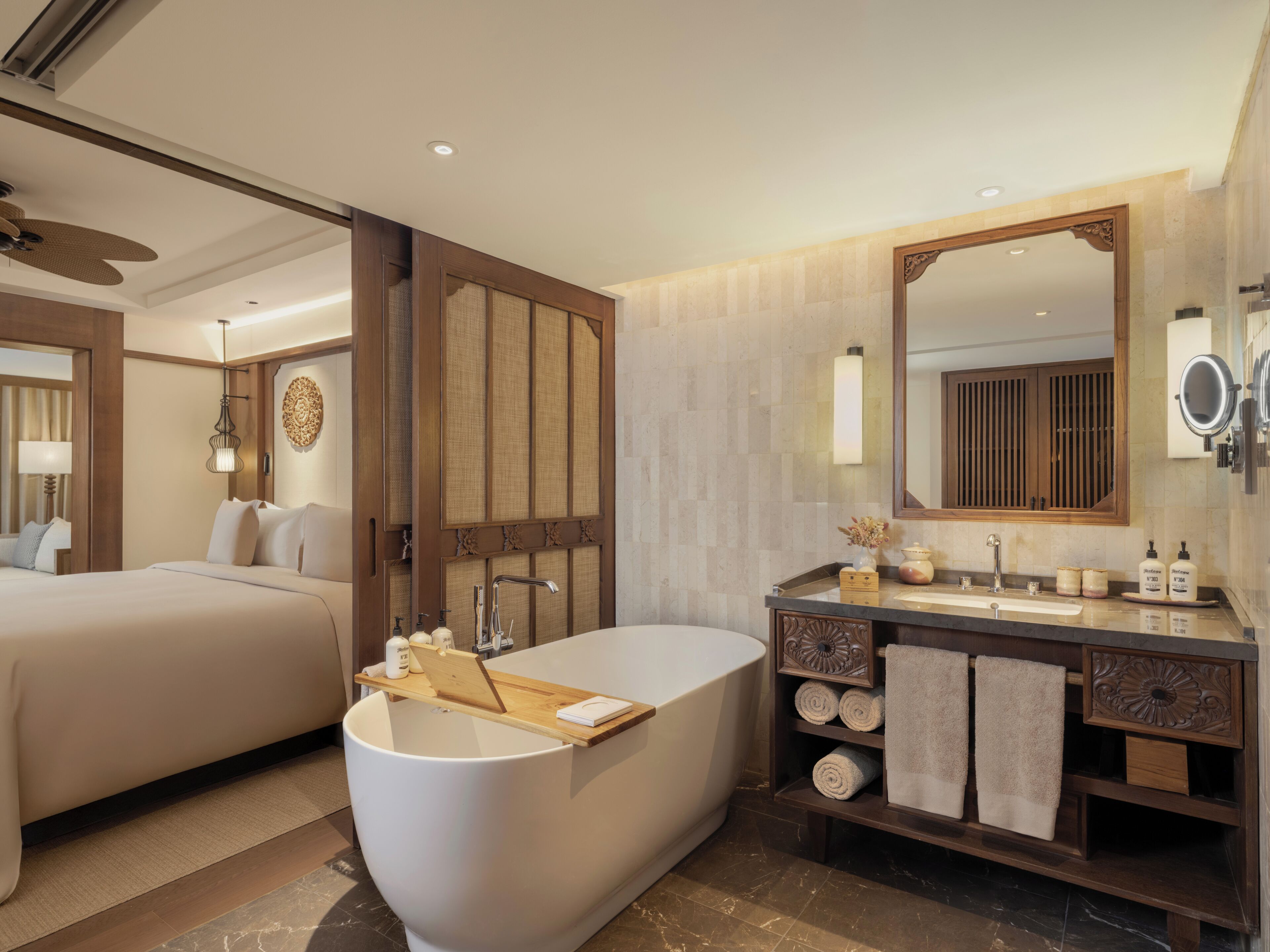 Uma casa de banho de hotel moderna com uma banheira independente, um lavatório com um espelho grande e um quarto separado com uma cama e uma mesa de cabeceira.