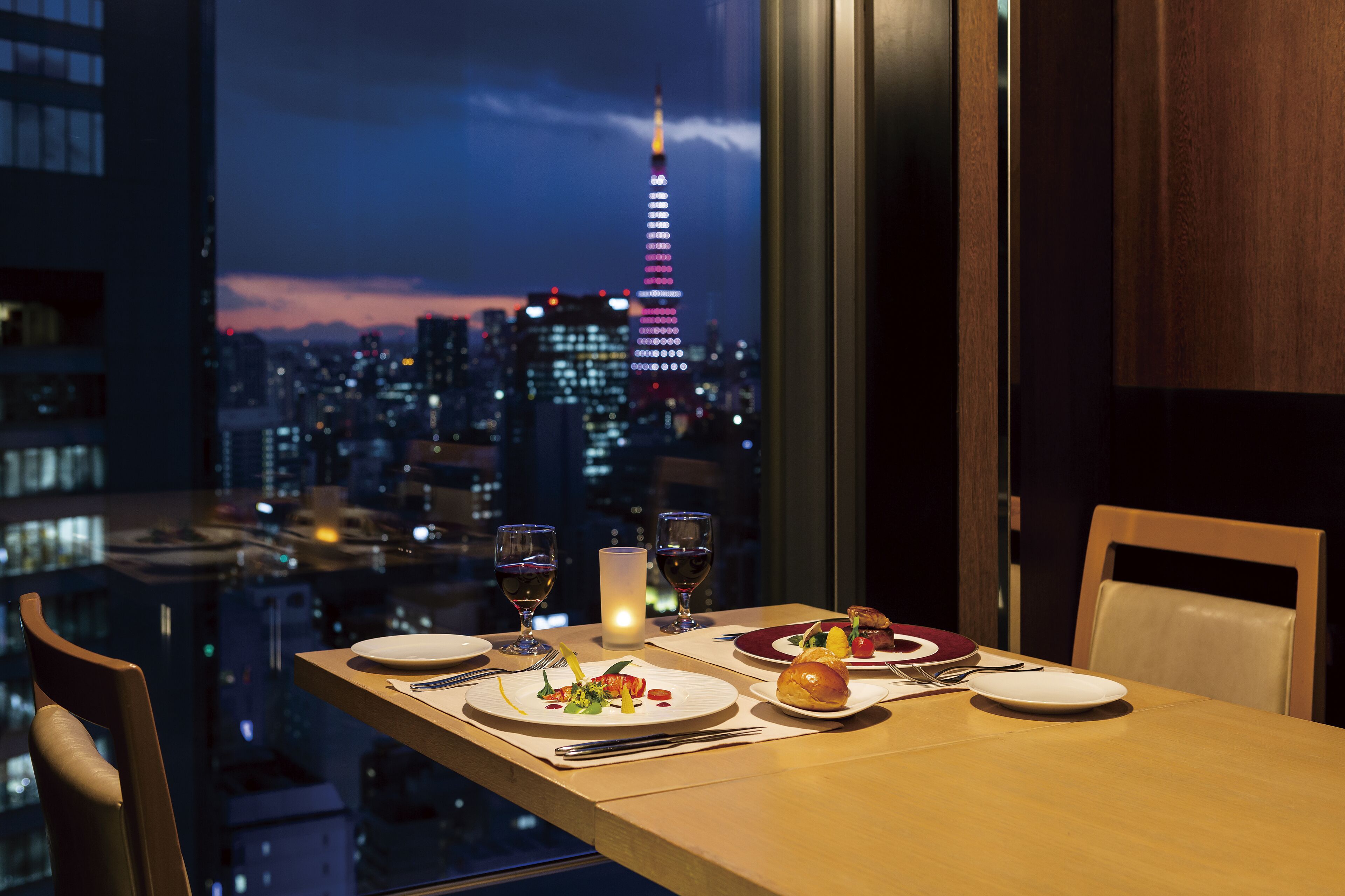 Mesa de jantar posta com pratos, copos e talheres, com vista para um horizonte urbano onde se destaca uma torre.