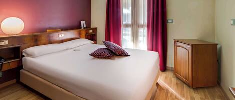 Quarto de hotel com uma cama grande, cabeceira de madeira, mesa de cabeceira com abajur, televisão fixada na parede e um pequeno armário de madeira.