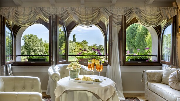 Quarto com vista para um jardim através de janelas em arco, uma mesa com lanches e bebidas, e um sofá.
