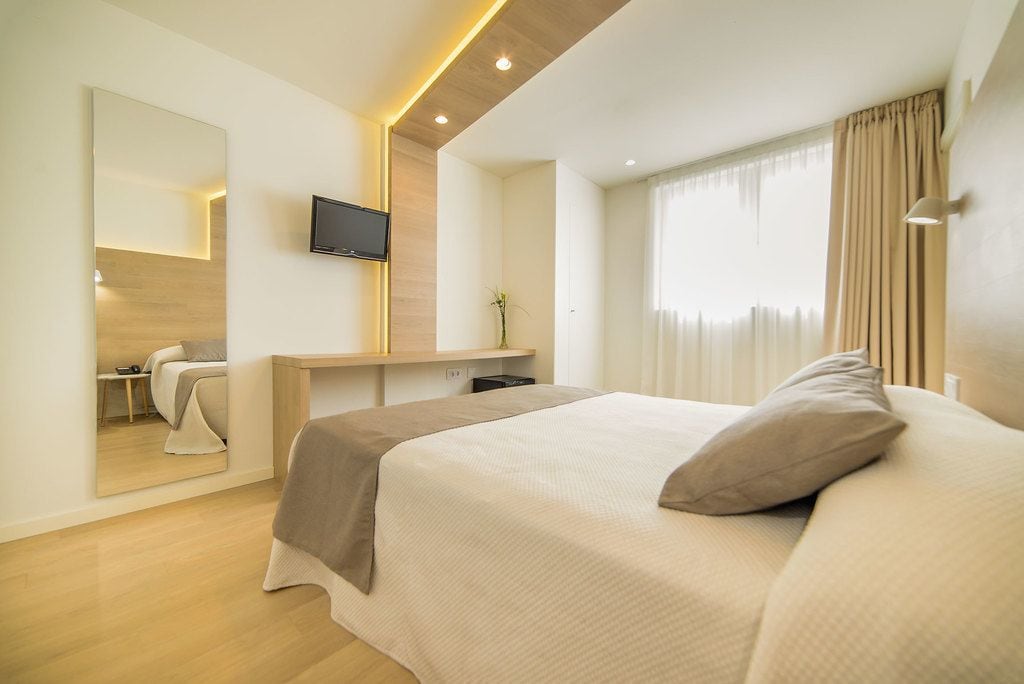 Quarto de hotel moderno com uma cama grande, televisão fixada na parede, uma escrivaninha e um espelho refletindo o quarto.