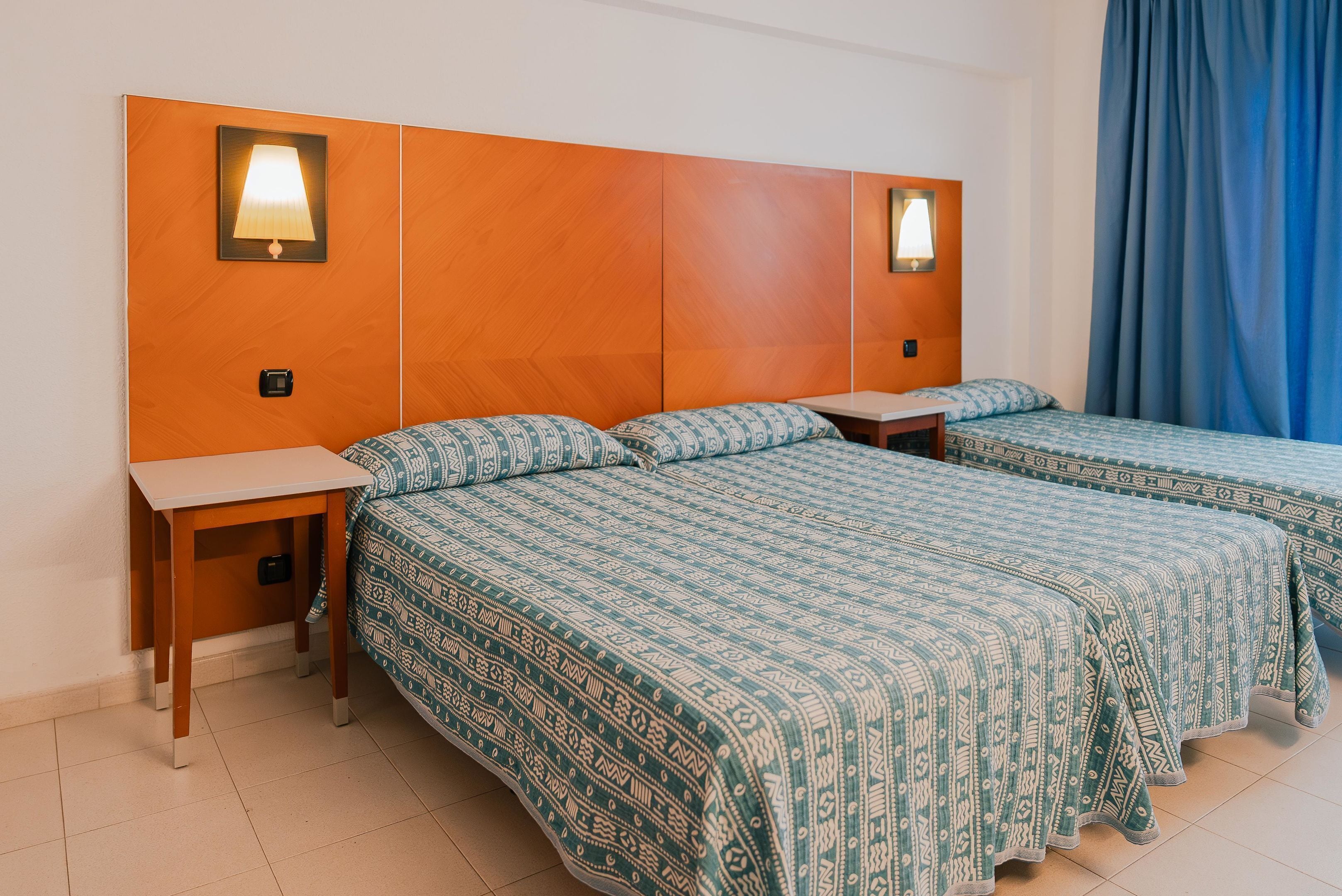 Quarto de hotel com duas camas, cada uma com roupa de cama estampada, mesinhas de cabeceira de madeira e luminárias fixadas na parede.