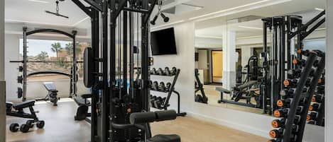 Uma academia com diversos equipamentos de exercício, um banco e uma TV fixada na parede.