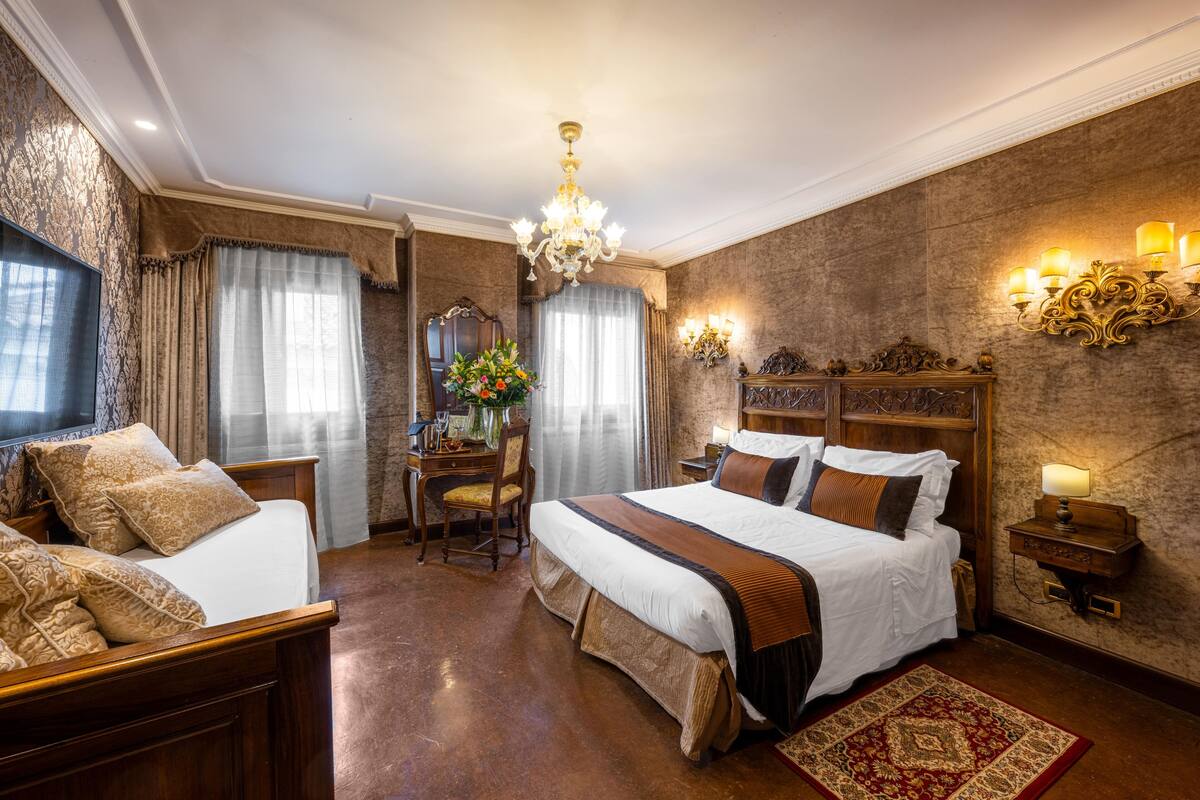 Quarto de hotel com uma cama grande, escrivaninha, cadeira, televisão e lustre.