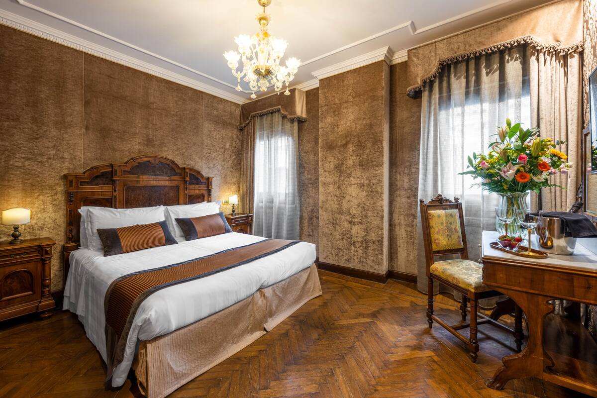 Um quarto espaçoso com uma cama grande, cabeceira de madeira, um candelabro, uma escrivaninha com cadeira e um vaso de flores.