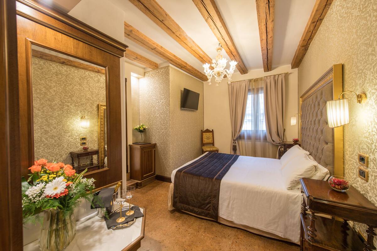 Quarto de hotel com uma cama grande, teto de madeira, lustre, televisão e um vaso de flores.