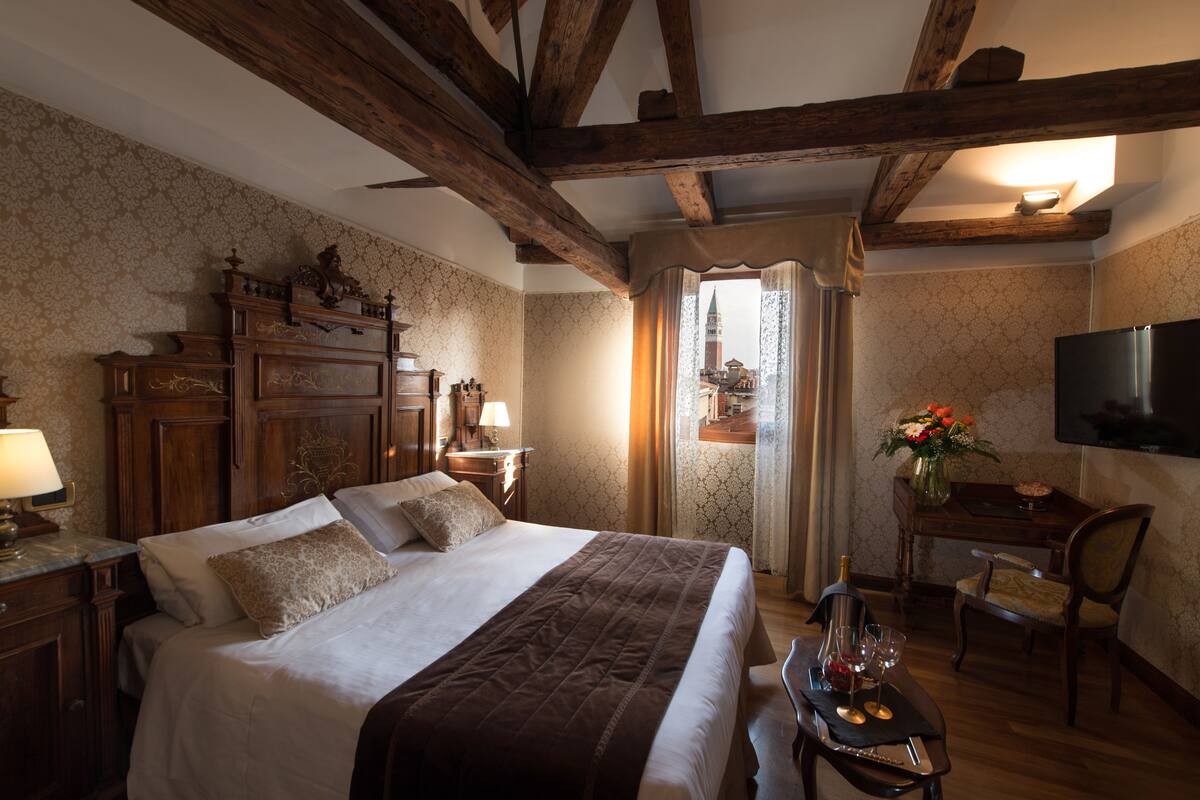 Quarto de hotel com uma cama grande, mobiliário de madeira, uma escrivaninha com cadeira, televisão e vista para uma torre pela janela.