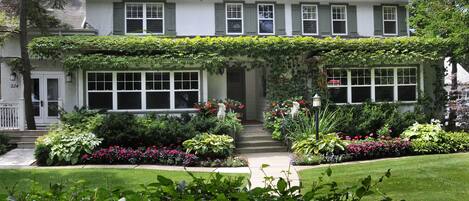 Uma casa de dois andares com varanda frontal, entrada coberta e um jardim bem cuidado com diversas plantas e flores.