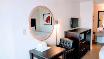 Quarto de hotel com cama, espelho redondo, escrivaninha com cadeira, cômoda, televisão e luminária.