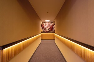 Um corredor de hotel com piso revestido de carpete, parede com padrão de folhagens e iluminação embutida ao longo das paredes.