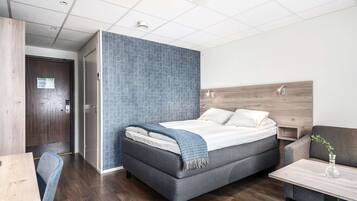 Quarto de hotel moderno com cama cinza, cabeceira de madeira, uma pequena mesa com um vaso e uma parede decorada com azulejos azuis.