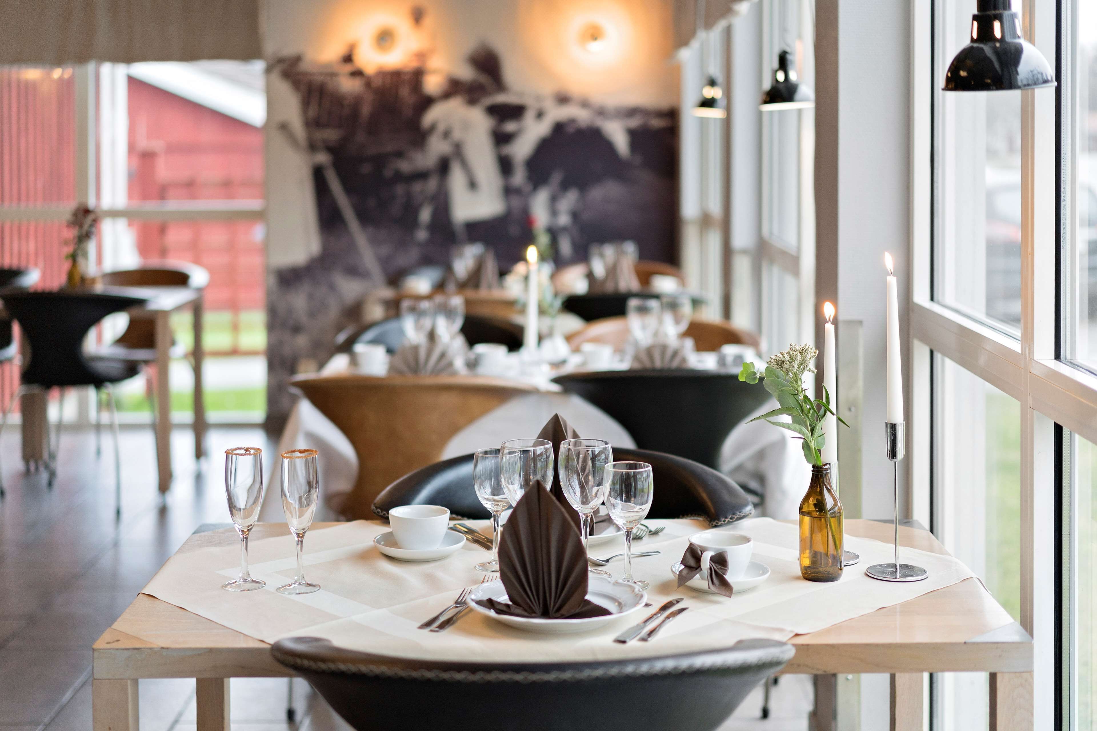 Uma sala de jantar com mesas preparadas para uma refeição, com velas, taças de vinho e um vaso com flores.