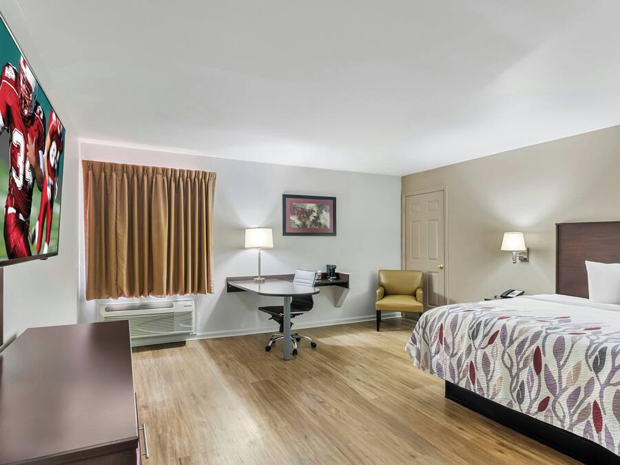 Quarto de hotel com cama, escrivaninha, cadeira, televisão e um quadro na parede.