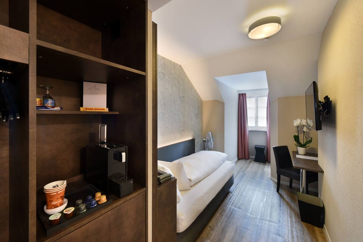 Quarto de hotel com cama, mesa de cabeceira, cadeira e uma pequena escrivaninha com uma planta em vaso.