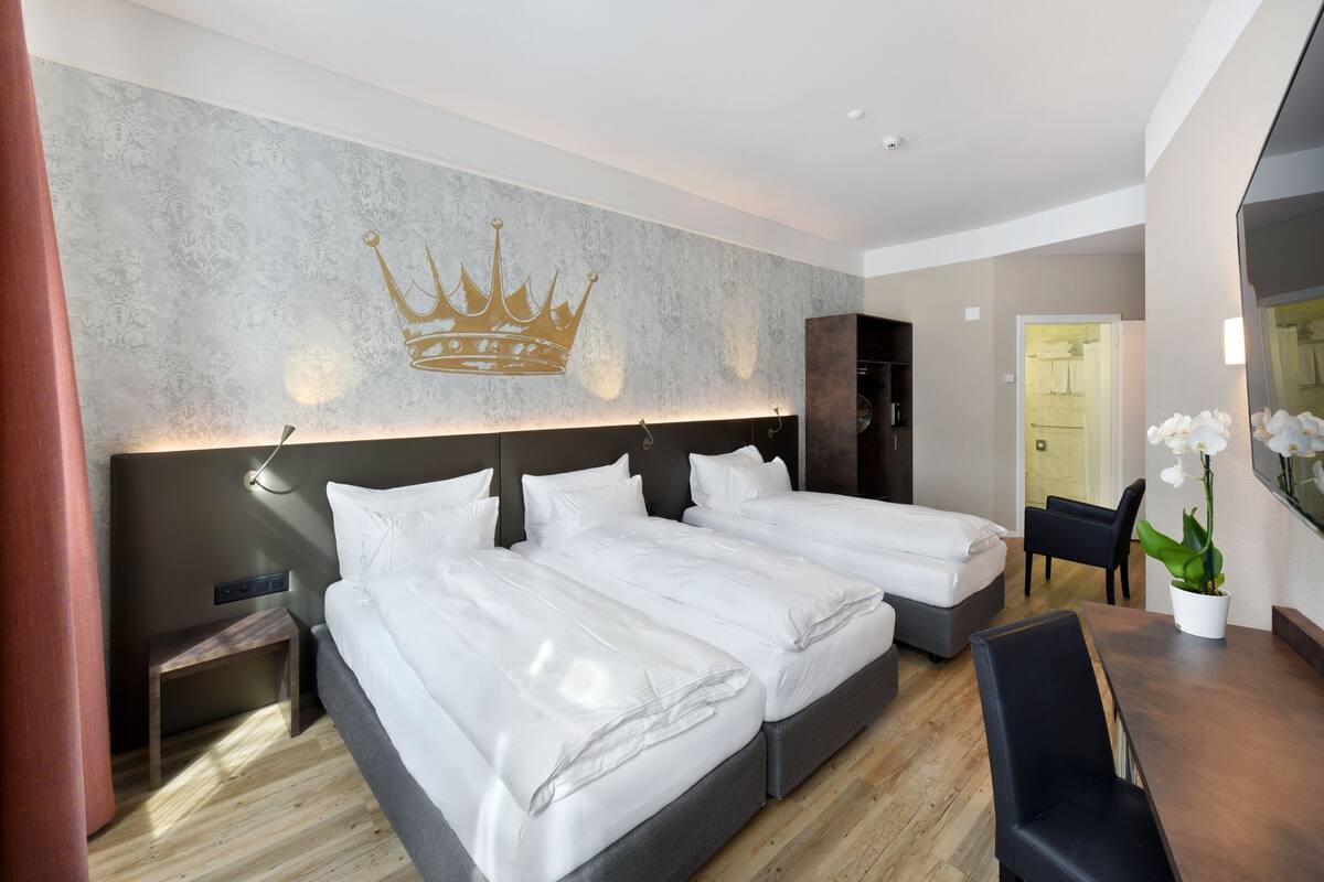 Quarto de hotel moderno com uma cama grande, uma mesa de cabeceira, uma escrivaninha pequena, uma cadeira, um vaso de planta e um espelho.