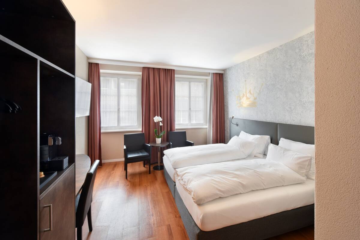 Quarto de hotel com cama, duas cadeiras, uma mesinha e escrivaninha com cafeteira.