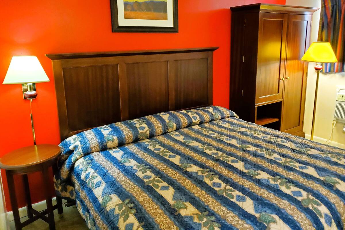 Quarto de hotel com uma cama, mesa de cabeceira, luminária, guarda-roupa e um quadro na parede.