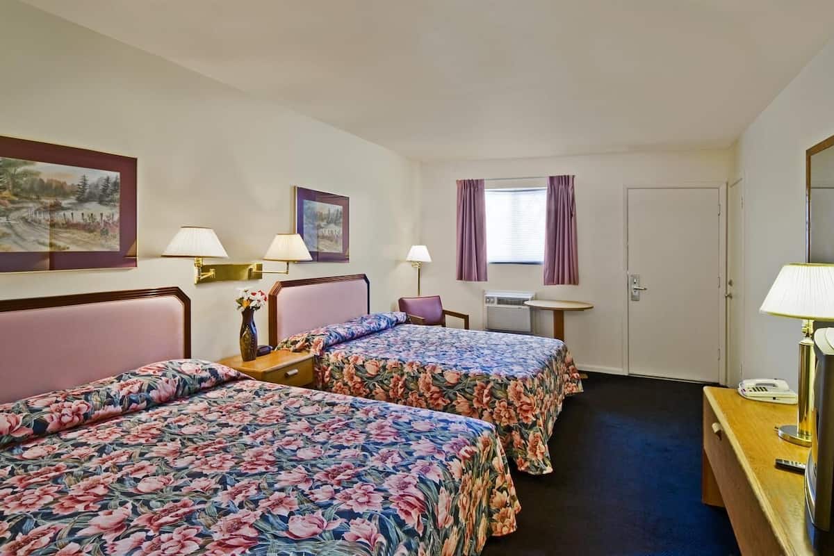 Quarto de hotel com duas camas, uma escrivaninha, uma cadeira, uma mesa pequena, um abajur, um quadro e uma janela com cortinas.