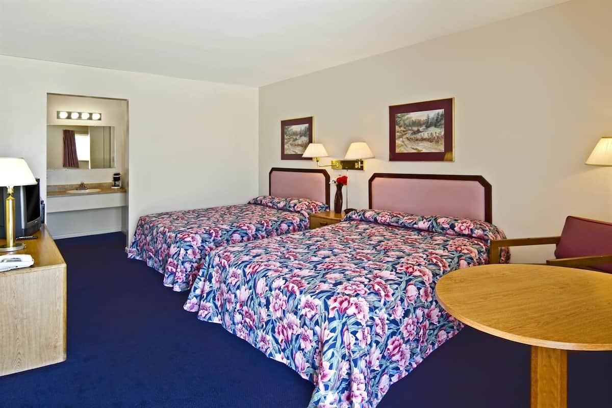 Quarto de hotel com duas camas, uma escrivaninha, uma cadeira, uma mesa redonda, um espelho com iluminação e duas luminárias de parede.