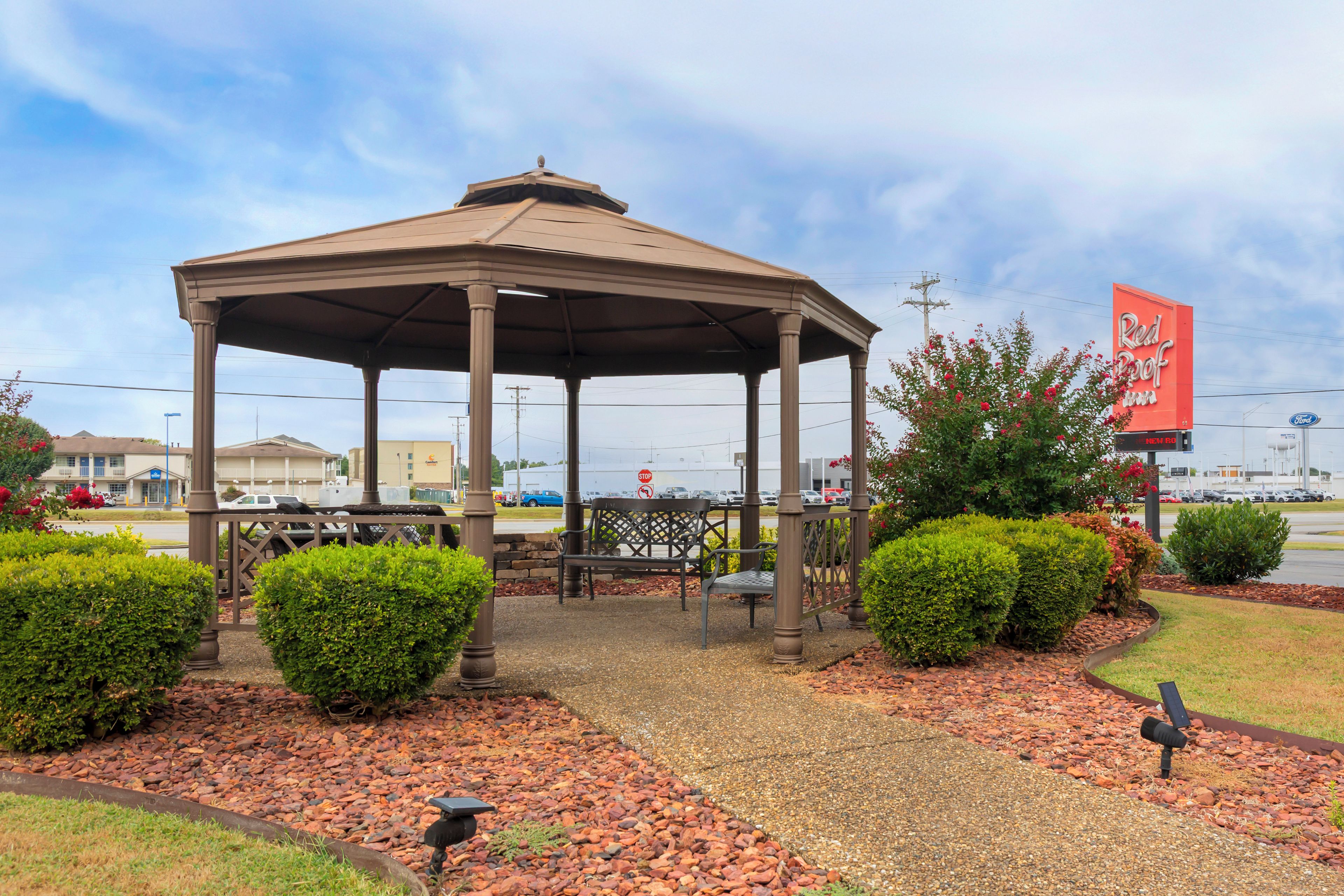 Um gazebo com um banco, rodeado por arbustos bem cuidados e um caminho de pedrinhas.